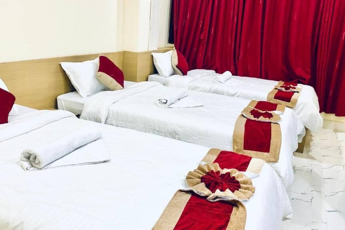 Quarto de hotel com três camas, cada uma com roupa de cama branca e detalhes vermelhos, cabeceira de madeira e cortinas vermelhas.