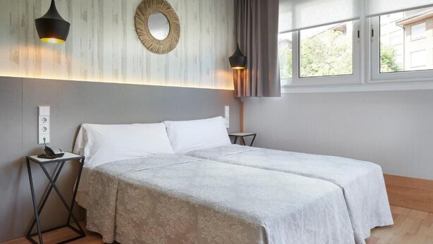 Uma cama de casal com colcha estampada, duas mesas de cabeceira, um espelho fixado na parede e uma janela com cortinas.