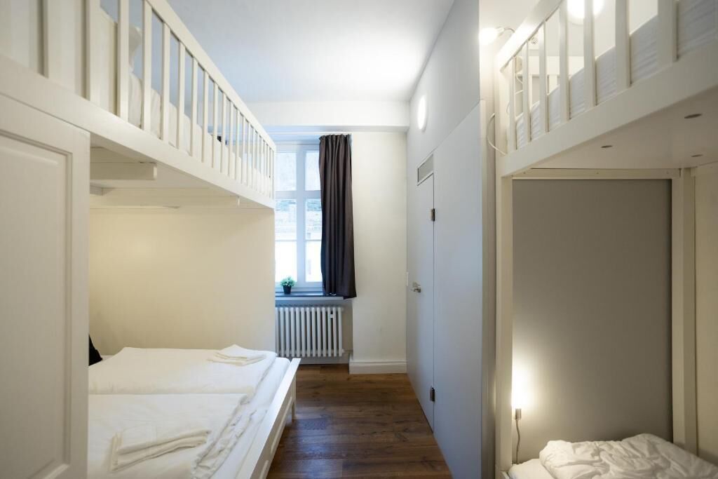 Quarto de hotel com beliche, janela com cortinas, radiador e porta.