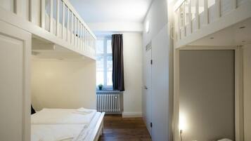 Quarto de hotel com beliche, janela com cortinas, radiador e porta.