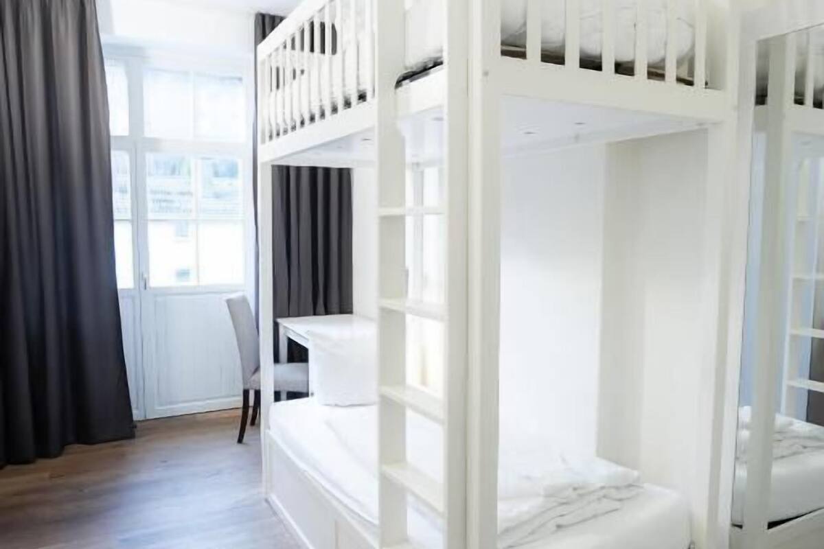 Quarto com beliche, escrivaninha e cadeira, piso de madeira e janela com cortinas.