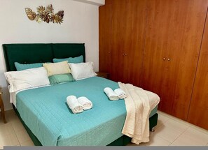 Uma cama bem arrumada com um edredom verde-água, duas toalhas enroladas e um cobertor bege.