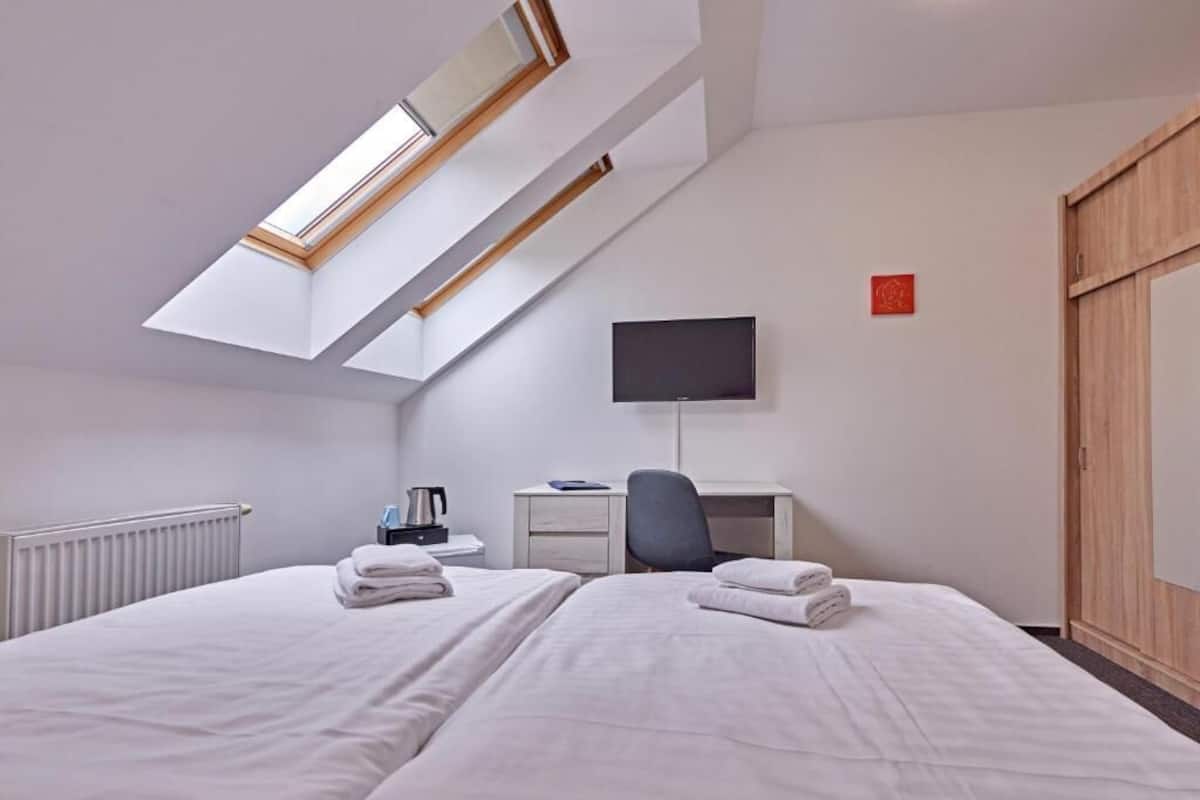 Quarto de hotel com duas camas, uma escrivaninha, uma cadeira, uma televisão e um claraboia.