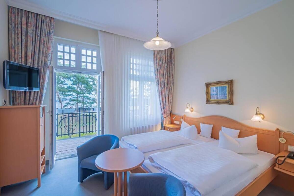 Quarto de hotel com duas camas, televisão, uma mesinha, uma cadeira e varanda com vista para árvores.