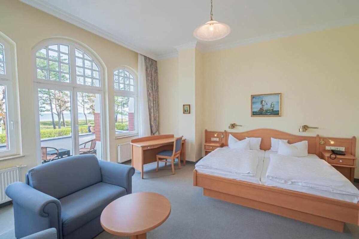 Quarto de hotel com cama, escrivaninha, poltrona, sofá e janelas amplas.