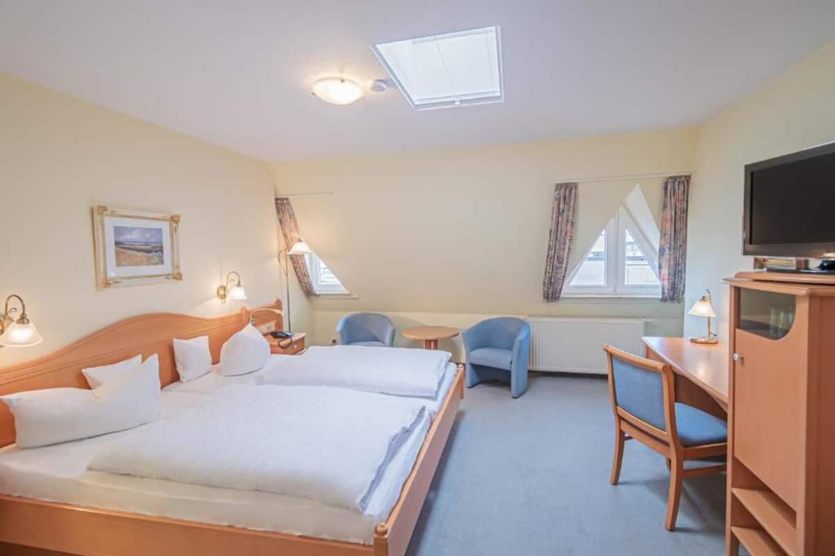 Quarto de hotel com uma cama de casal, uma escrivaninha, uma cadeira, uma televisão e uma janela com cortinas.