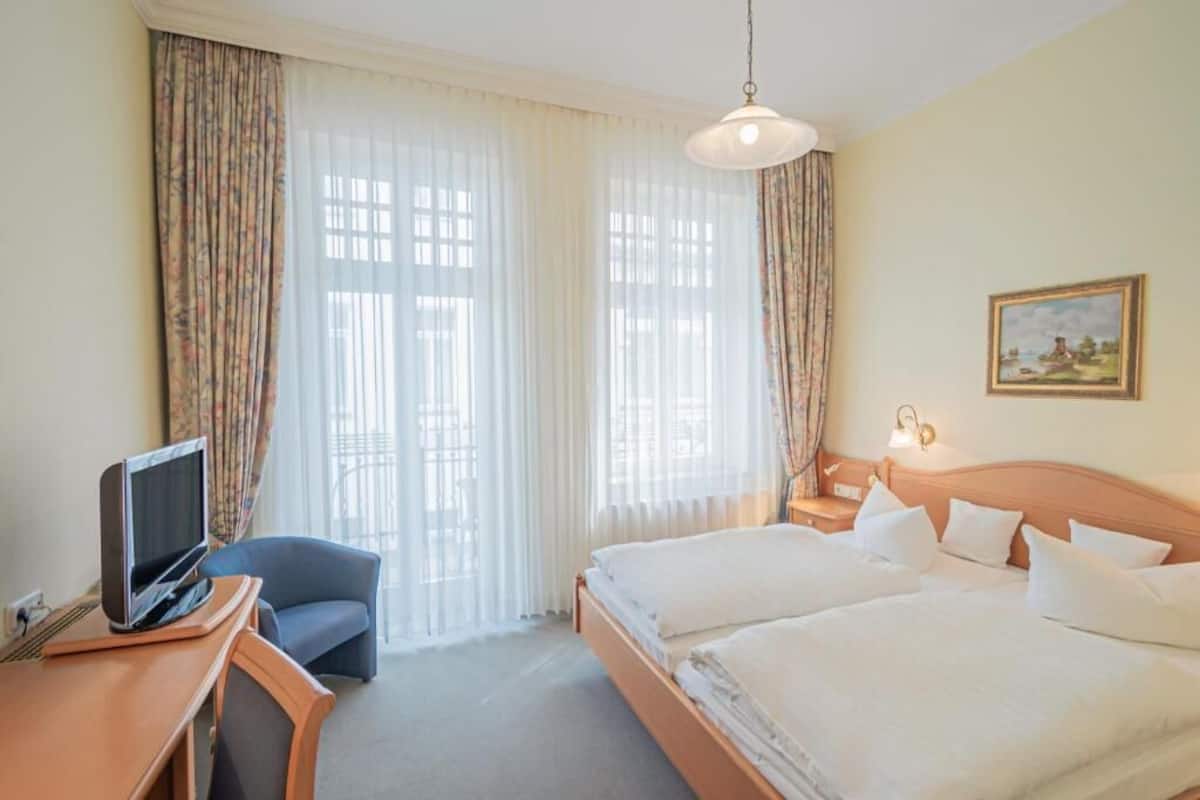 Quarto de hotel com cama, escrivaninha com televisão, cadeira e um quadro na parede.