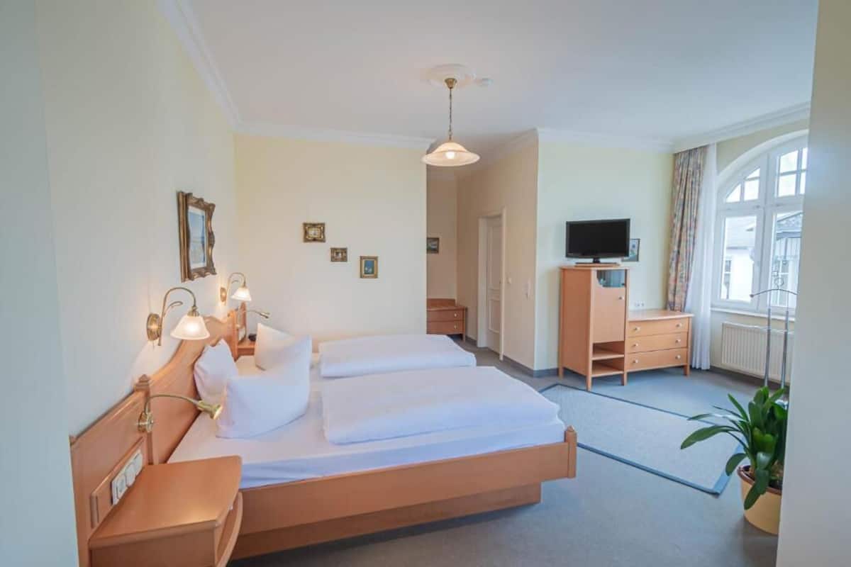 Quarto de hotel com cama, mesa de cabeceira, luminária, televisão, guarda-roupa e janela.