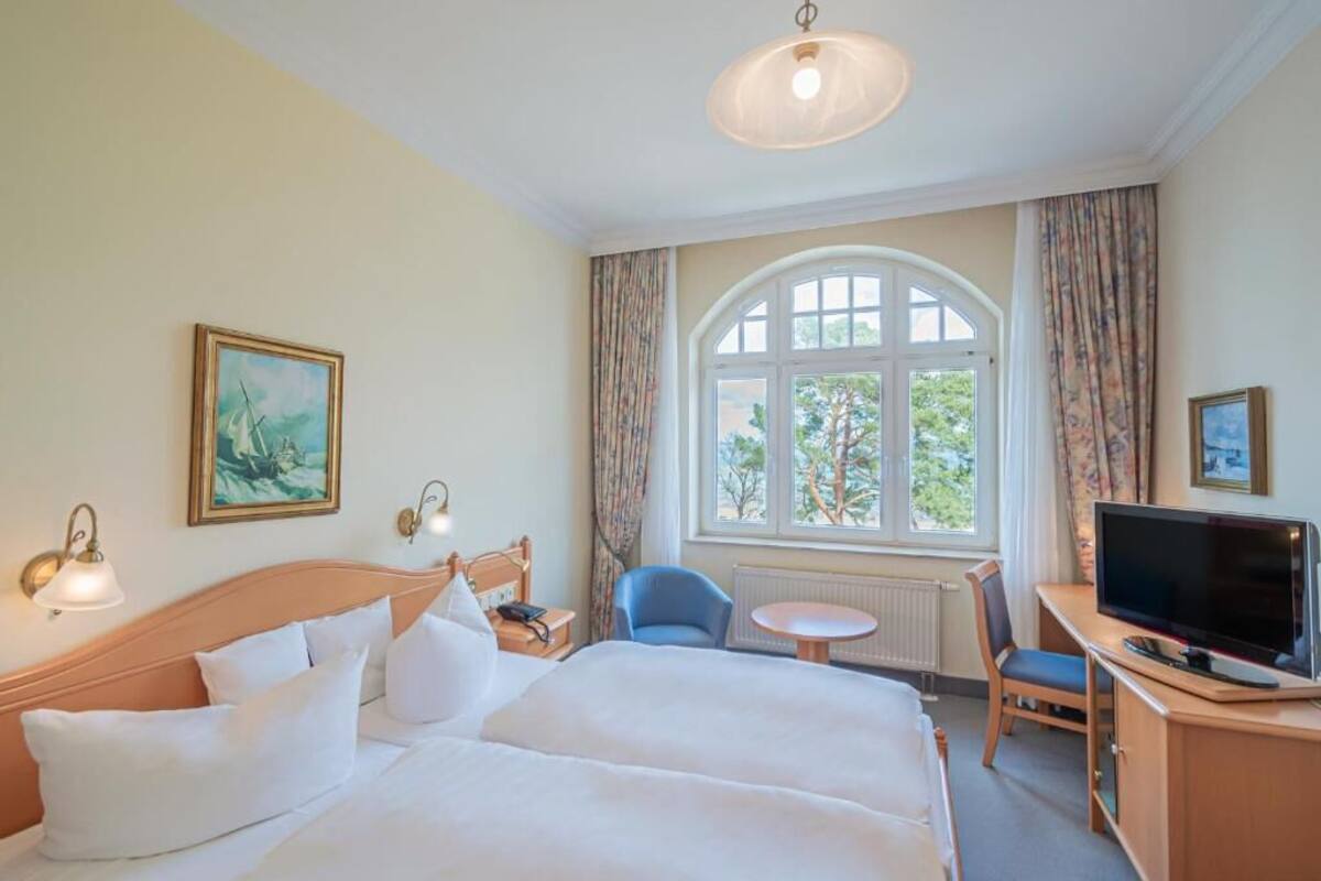 Quarto de hotel com cama, mesa de trabalho com televisão, cadeira, mesinha e janela com cortinas.