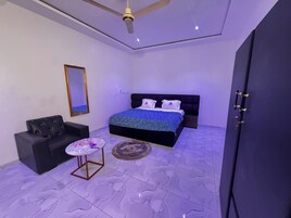 Quarto com uma cama, uma cadeira, uma mesinha, um espelho e um ventilador de teto.