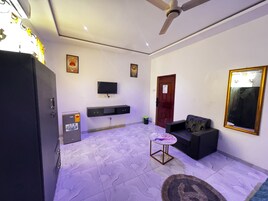 Quarto com ventoinha no teto, televisão fixada na parede, sofá, mesa redonda pequena e frigorífico.