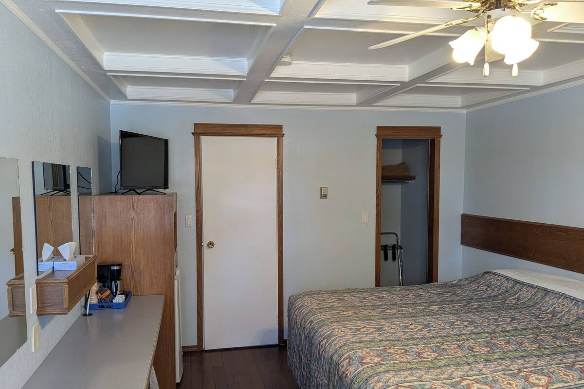 Um quarto com cama, escrivaninha, espelho, televisão, ventilador de teto e porta.