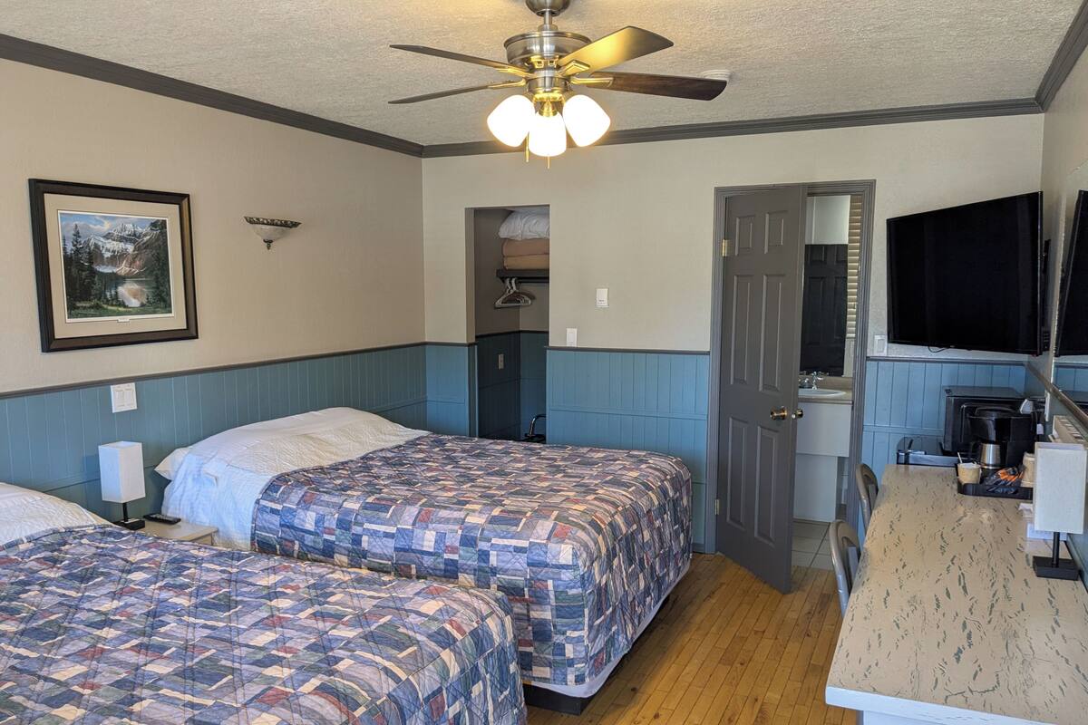 Quarto com duas camas, um ventilador de teto, um quadro emoldurado, uma porta que dá para uma cozinha e uma mesa com cafeteira.