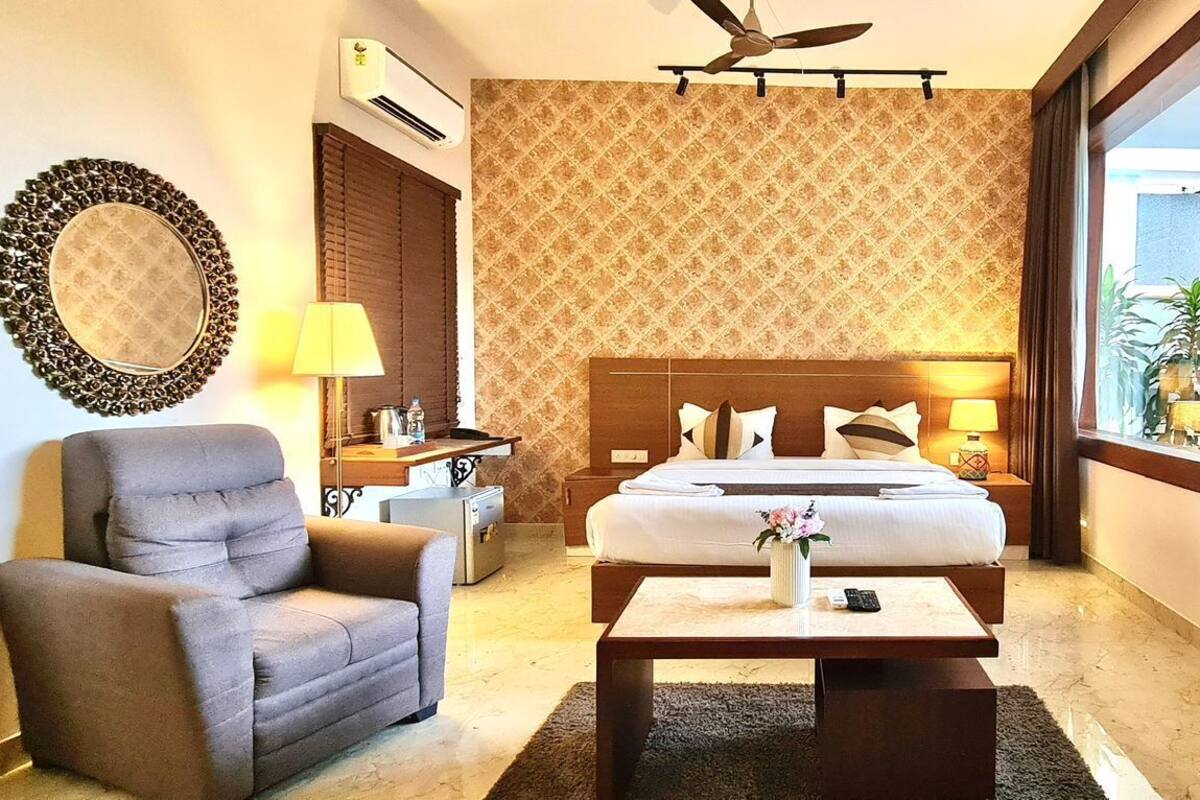 Quarto de hotel com cama, uma cadeira, escrivaninha, espelho e janela com vista para palmeiras.