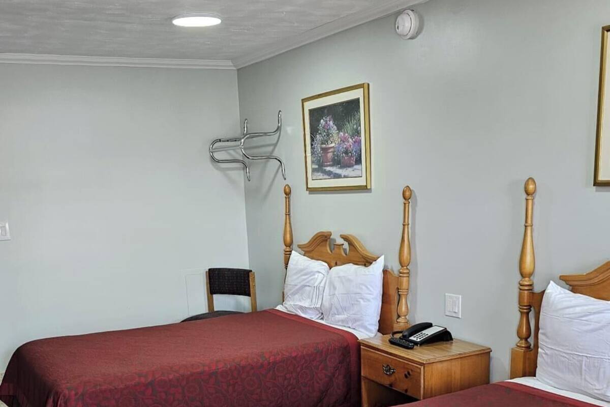 Quarto de hotel com duas camas, cabeceira de madeira, pequena mesa de cabeceira com telefone, ar condicionado fixado na parede e quadro emoldurado.