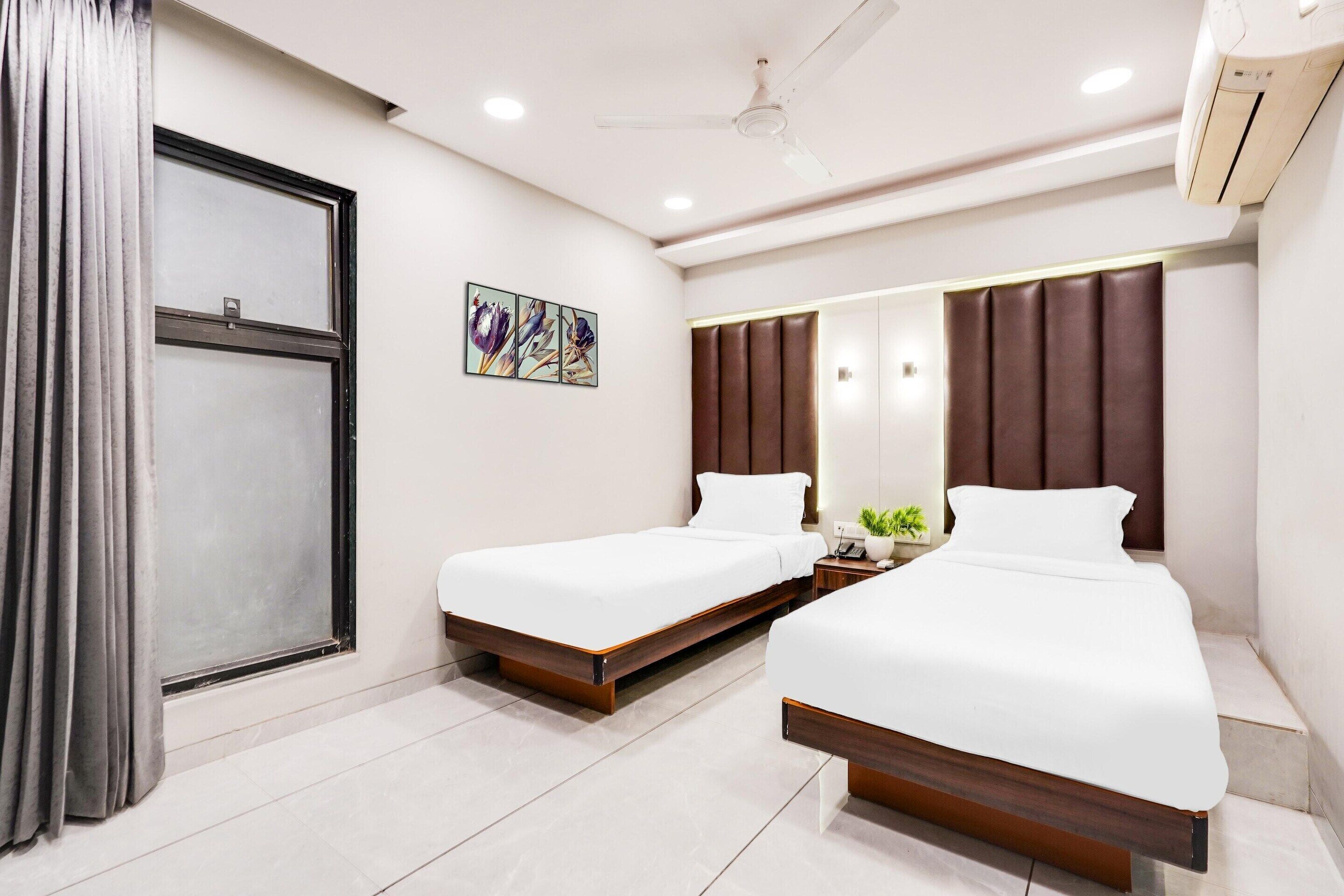 Quarto de hotel com duas camas, uma janela grande com cortinas, um ventilador de teto, iluminação embutida e ar condicionado fixado na parede.