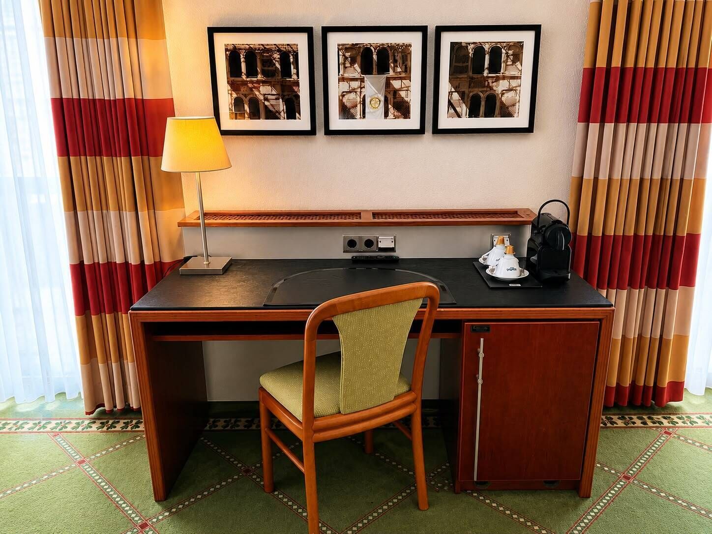 Quarto de hotel com escrivaninha, cadeira, luminária e três quadros na parede.