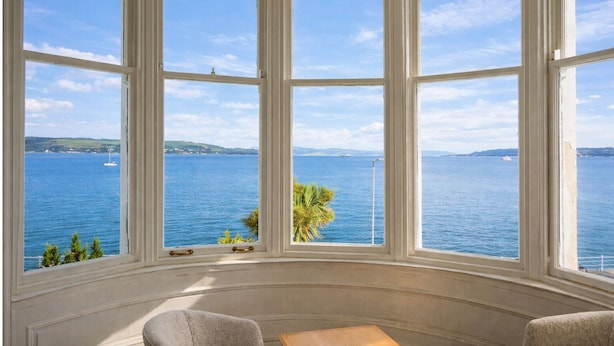 Quarto com janela em sacada com vista para a água, uma cadeira e uma pequena mesa.