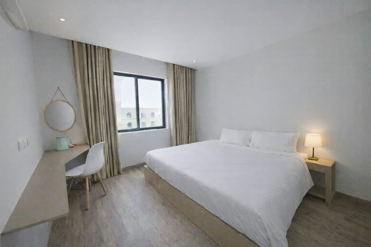 Quarto de hotel com uma cama, escrivaninha com cadeira, luminária e janela com cortinas.