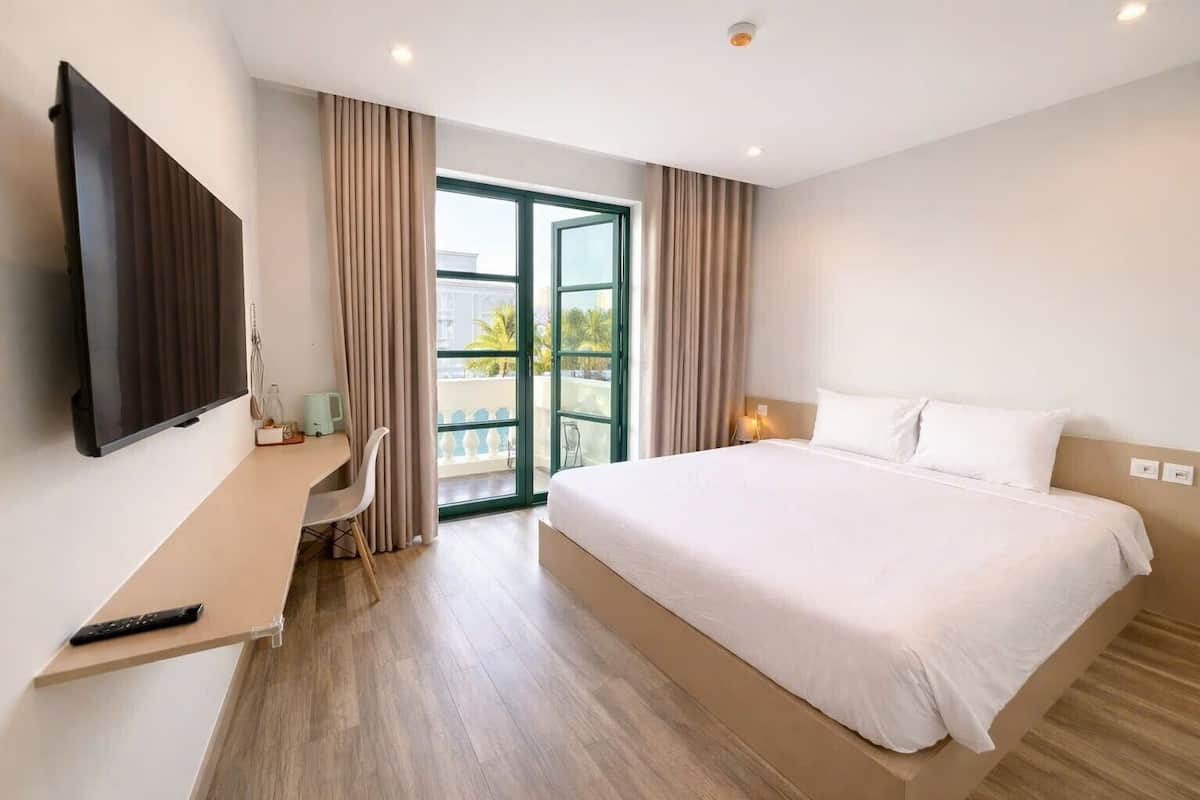 Quarto de hotel com uma cama, escrivaninha, televisão e varanda com cortinas.