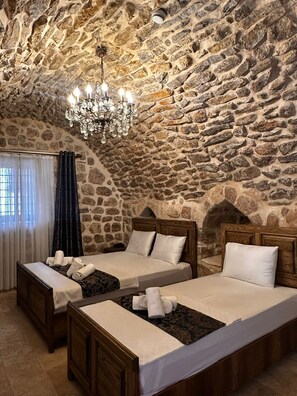 Quarto com parede de pedra, um candelabro, duas camas com cabeceiras de madeira e uma janela com cortinas.