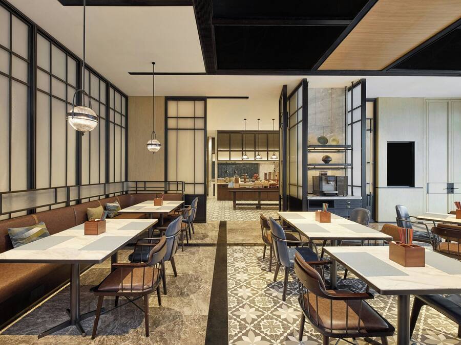 Restaurante moderno com mesas altas, bancos e uma cozinha ao fundo, além de um piso com padrão.