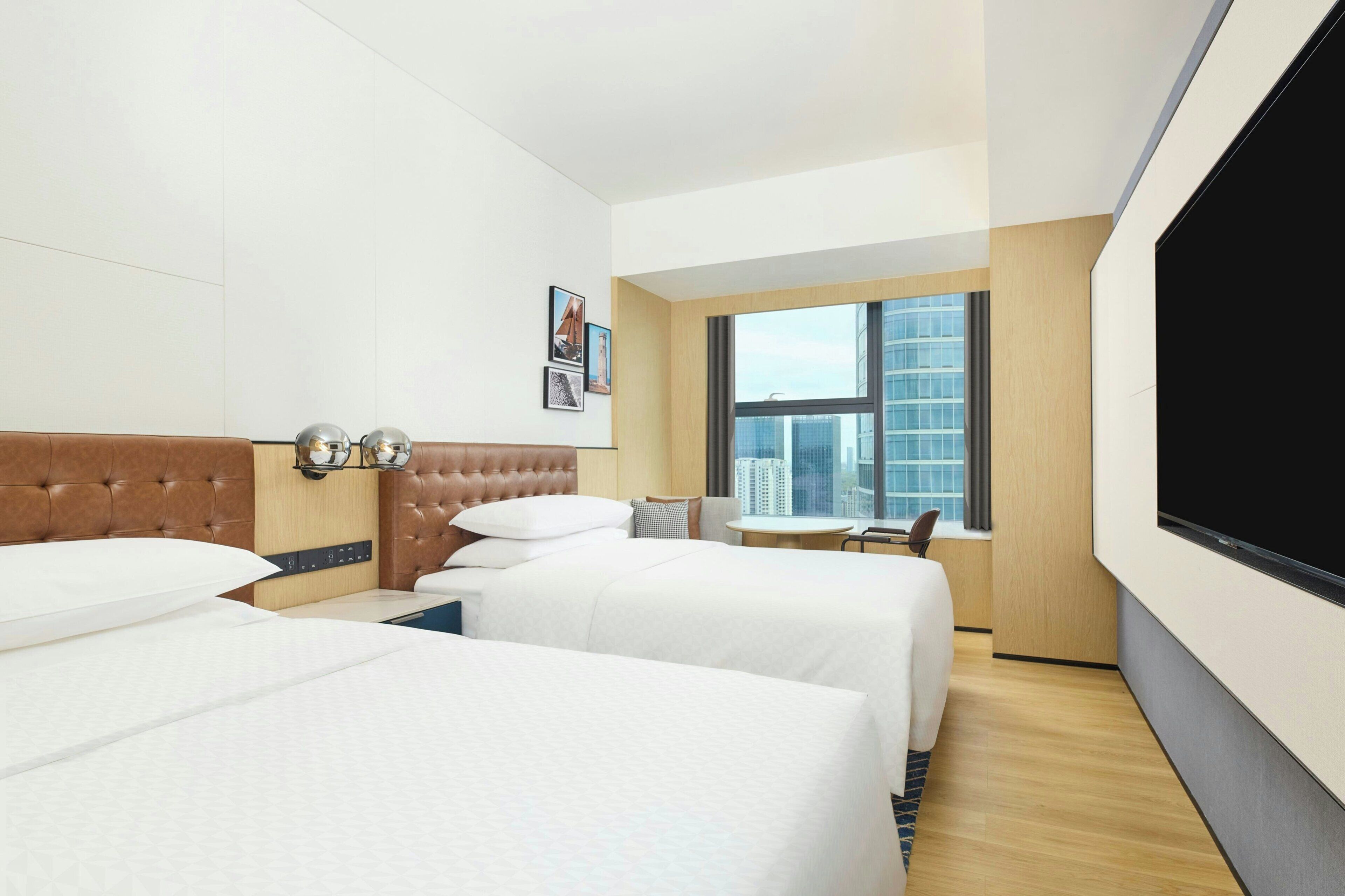 Quarto de hotel com duas camas, televisão, janela com vista para a cidade e uma pequena mesa.