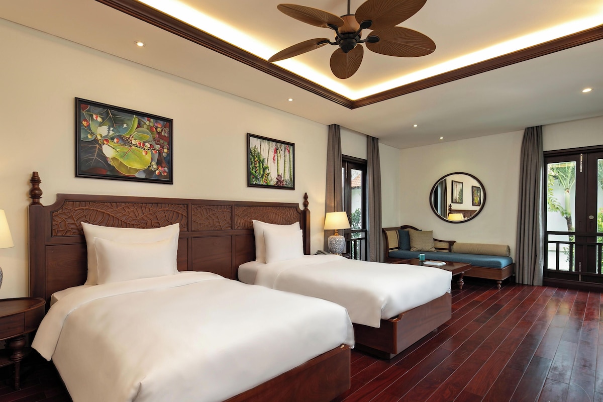 Quarto de hotel com duas camas, ventoinha no teto, obras de arte nas paredes, um sofá e vista pela janela.