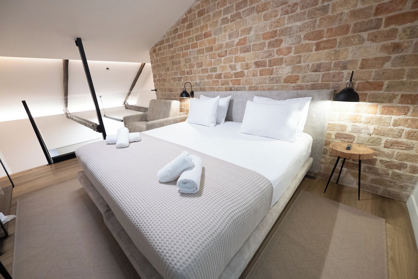 Quarto moderno com cama grande, mesa de cabeceira, cadeira e parede de tijolos.