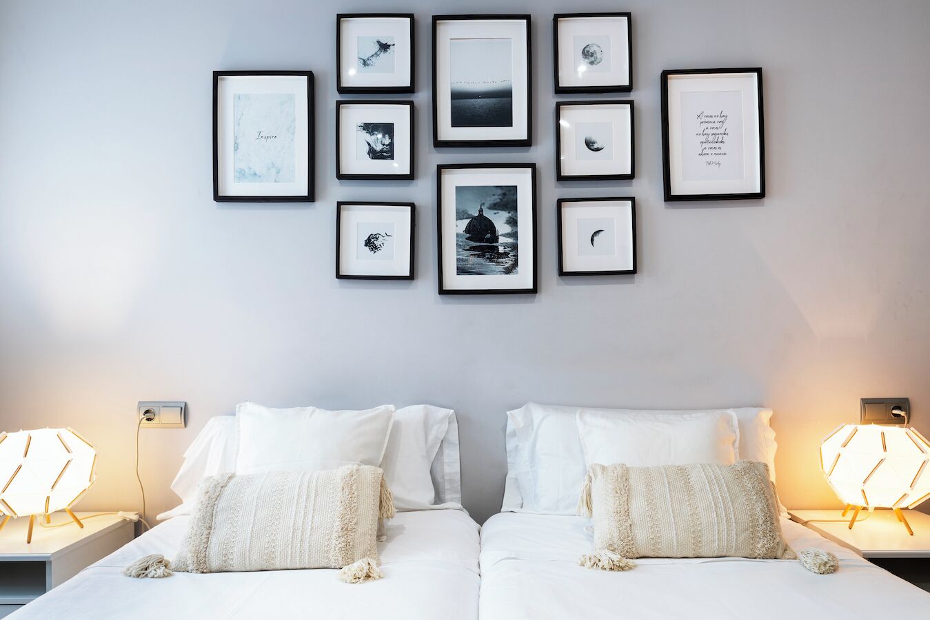 Uma cama de casal com lençóis brancos e duas almofadas decorativas, uma parede com nove quadros emoldurados e duas luminárias de cabeceira.