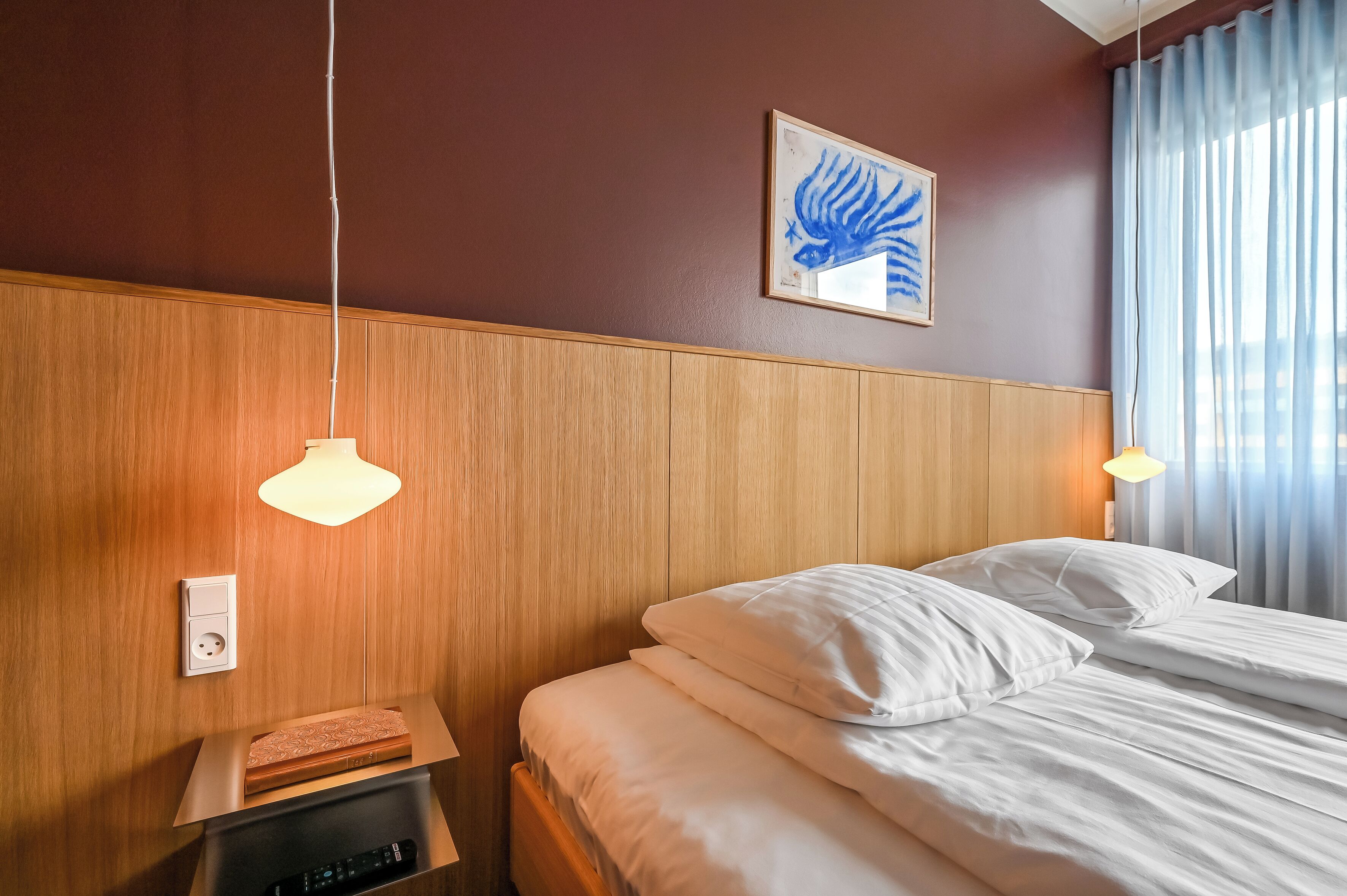 Quarto de hotel com cabeceira de madeira, cama com lençóis brancos, mesa de cabeceira com controle remoto e luminária fixada na parede.