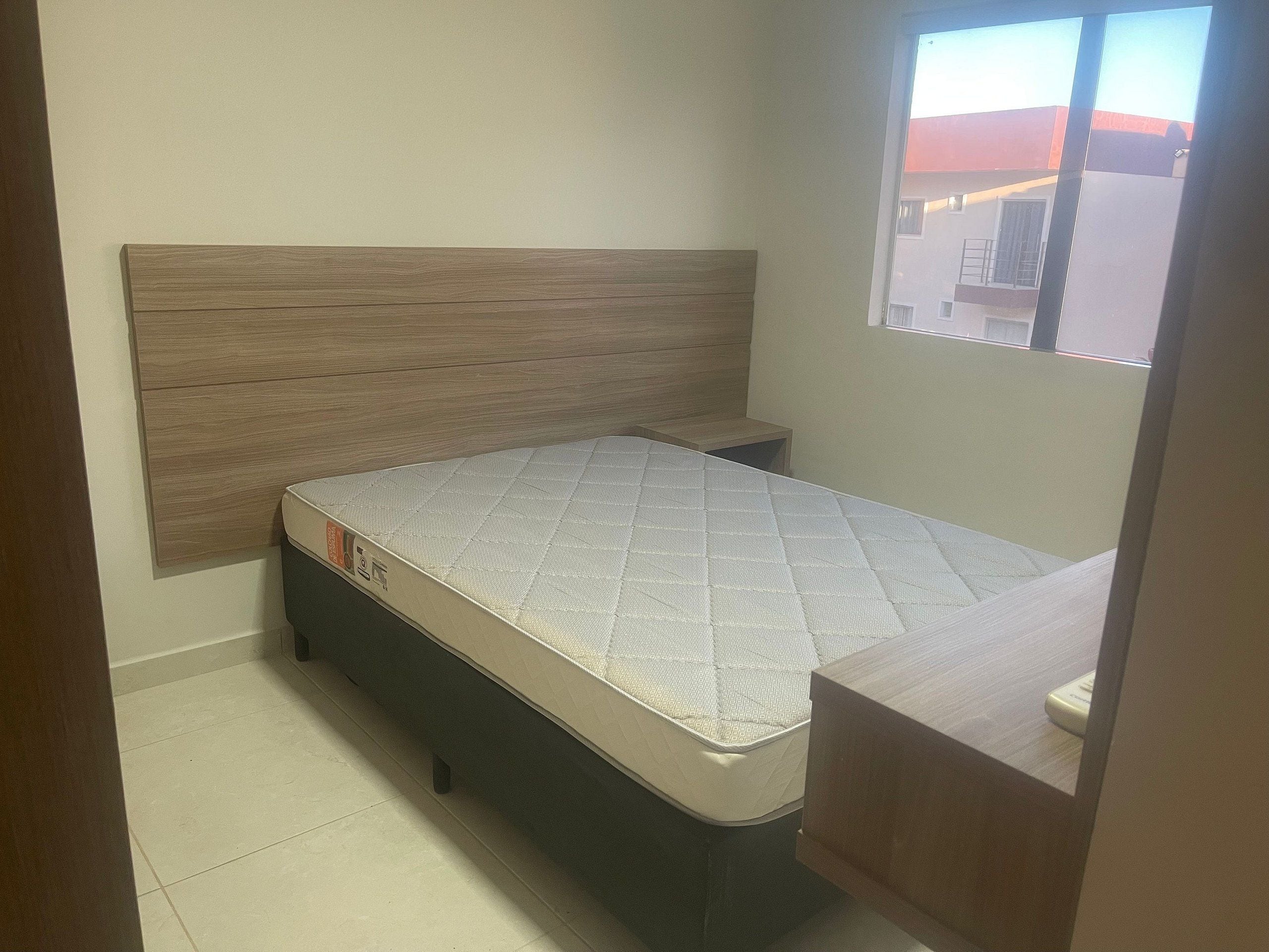 Um quarto com cama, cabeceira de madeira, uma mesinha de cabeceira e uma janela.