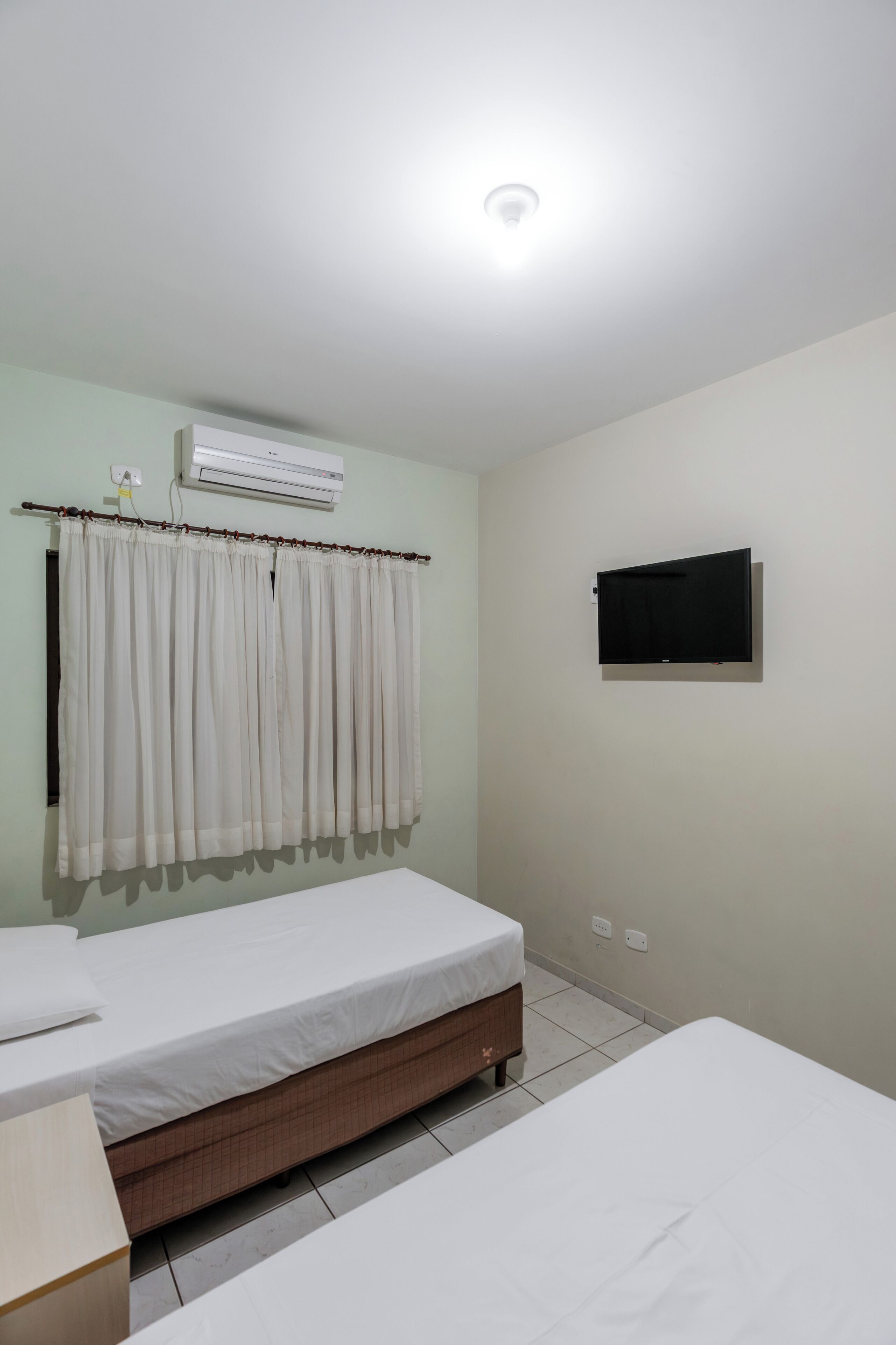 Quarto com duas camas, TV fixada na parede e aparelho de ar condicionado.
