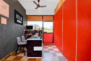 Uma receção de hotel com balcão de check-in, duas cadeiras, um ventilador de teto e uma janela grande com vista para edifícios e árvores.