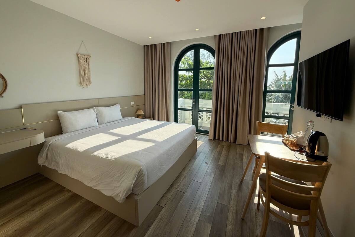 Quarto de hotel com cama, escrivaninha, cadeira, televisão e amplas janelas.