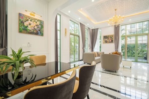 Uma sala de estar espaçosa com uma mesa de centro de vidro, assentos confortáveis, janelas amplas e um candelabro.