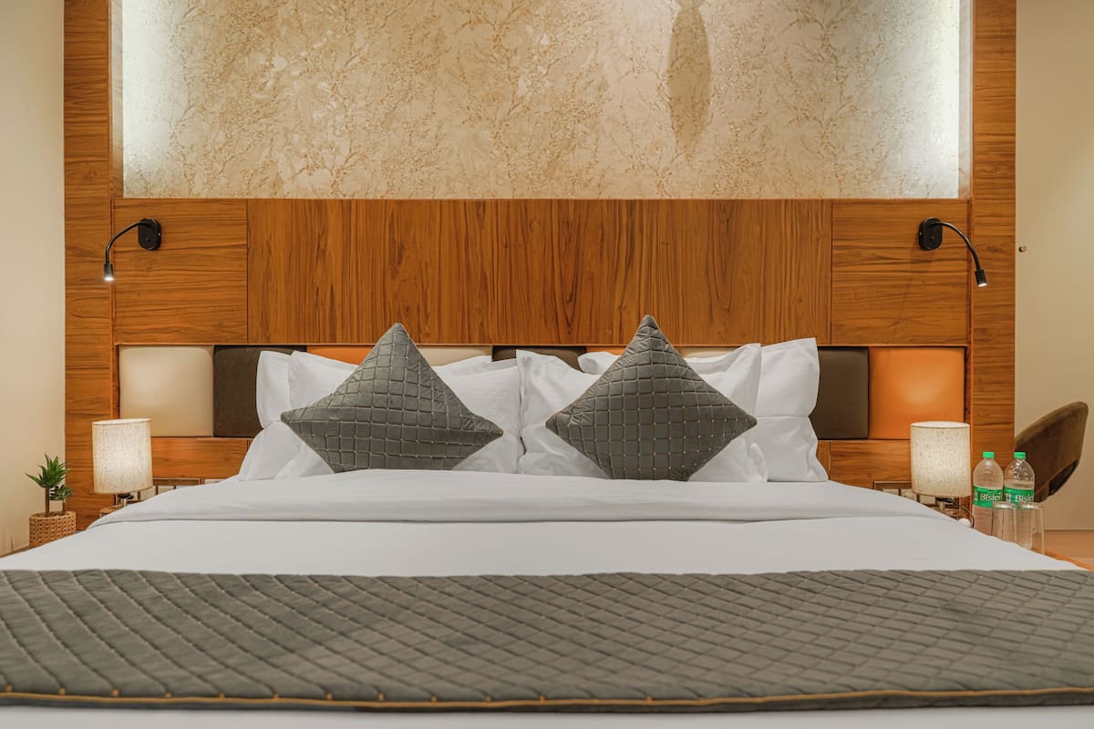 Quarto de hotel com cabeceira de madeira, duas luminárias de apoio, cama com roupa de cama branca e duas almofadas.