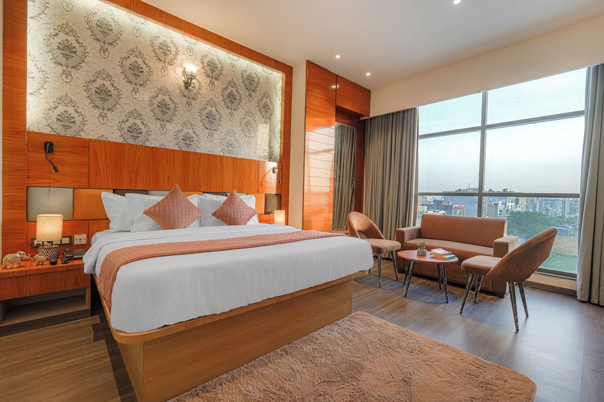 Quarto de hotel com uma cama grande, um sofá, uma pequena mesa e vista da cidade através de amplas janelas.