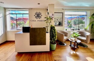 Balcão de recepção com logotipo, planta em vaso, sofá e vista de edifícios através de grandes janelas.