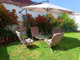 Um pátio externo com mesa e cadeiras, cercado por plantas em vasos e uma parede branca.