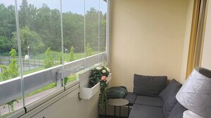 Uma varanda com sofá, uma mesinha, abajur e vista para o verde.