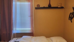 Uma cama de solteiro com colchão, mesa de cabeceira, janela com cortinas, prateleira com objetos decorativos e um sofá.