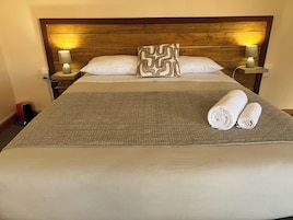 Quarto de hotel com cabeceira de madeira, duas luminárias de apoio, almofada estampada, toalha enrolada e colcha texturizada.