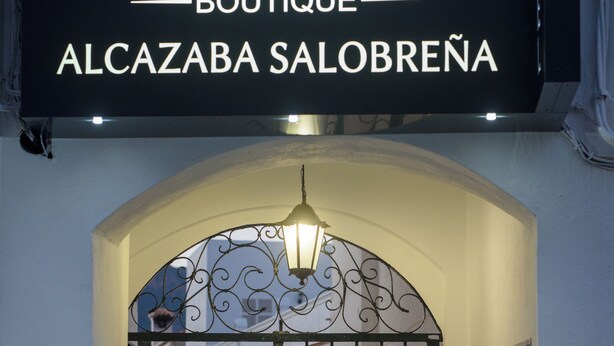 Entrada de hotel com portão de ferro forjado, uma placa indicando 'HOTEL BOUTIQUE ALCAZABA SALOBREÑA' e um caminho que leva ao interior.