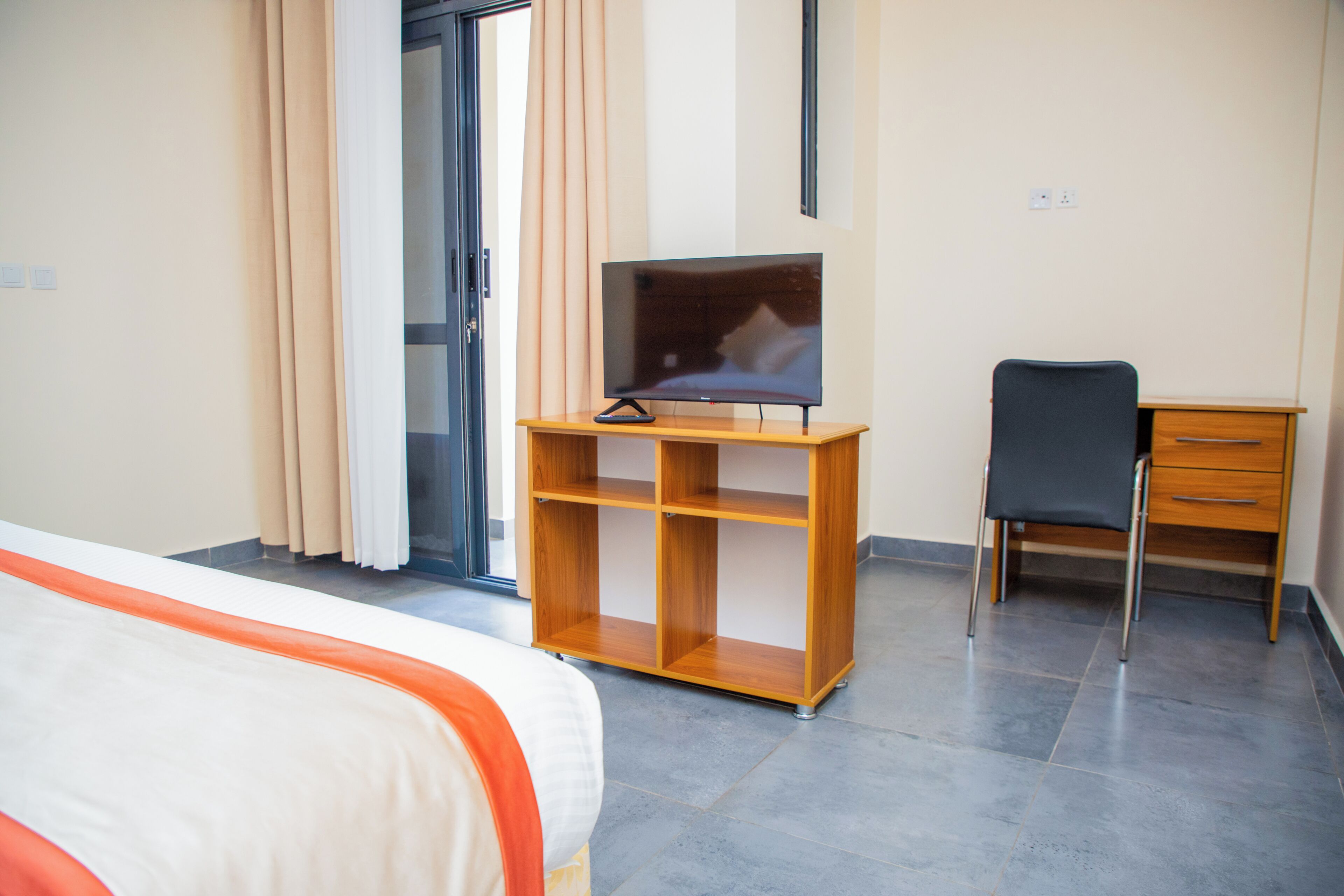 Quarto de hotel com cama, televisão sobre um suporte de madeira, escrivaninha com cadeira e porta de vidro deslizante.