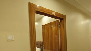 Um corredor com uma porta de madeira com a placa '104', dando acesso a um quarto com uma cama e um travesseiro branco.
