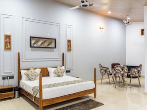 Quarto com cama de madeira, uma pequena mesa de cabeceira, um cesto de lixo, um ventilador de teto e um ponto de luz fixado na parede.