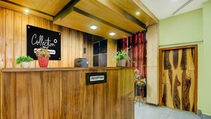 Balcão de recepção com uma placa que diz 'Collection' e um letreiro 'RECEPTION' abaixo. Há plantas em vasos no balcão, e uma porta com design de painel de madeira é visível ao fundo.