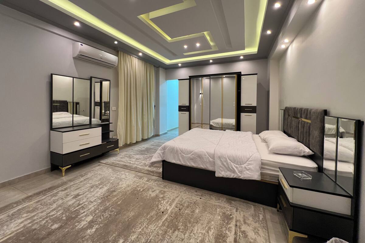 Quarto moderno com cama grande, mesa de cabeceira, guarda-roupa e espelho.