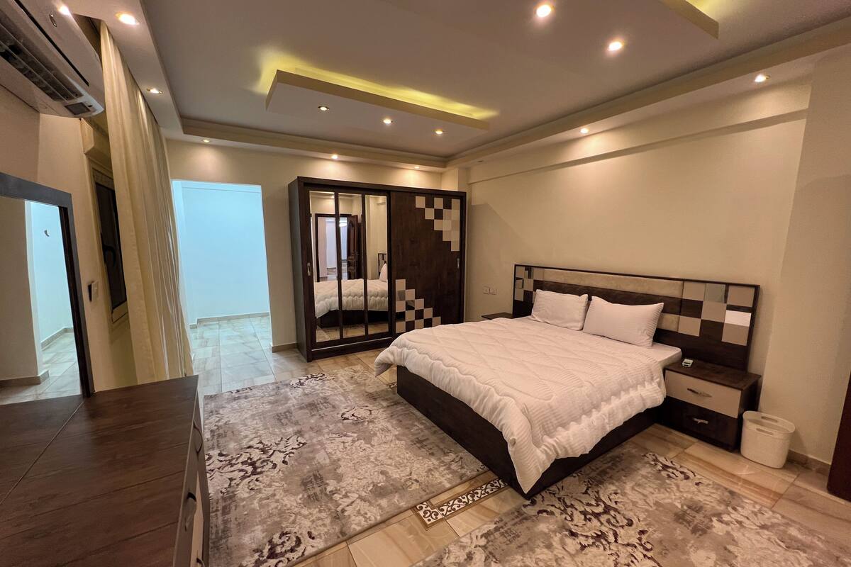 Quarto moderno com cama grande, mesas de cabeceira, guarda-roupa e tapete estampado.